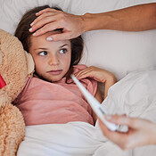 Sick little girl in bed with her teddybear while her mother uses a thermometer to check her ...