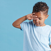 Sad little hispanic boy looking sad and rubbing his eyes against a blue ...